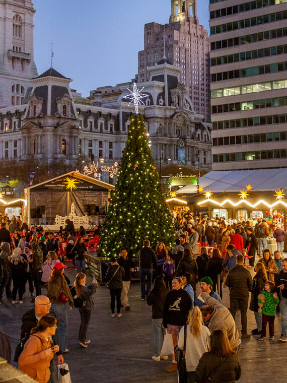 Vendor huts with decorative lighting and a large Christmas tree are set up in Love Park for Philadelphia's Christmas Village. People walk around shopping and taking photos. City Hall is in the background.