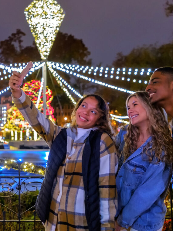 Three young adults wearing jackets and scarfs pose for a photo in front of a display of string lights at Franklin Square.