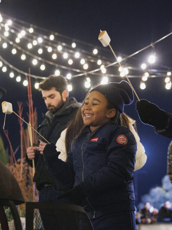 Children wearing winter jackets, hats and gloves roast marshmallows over a fire pit under a canopy of string lights.