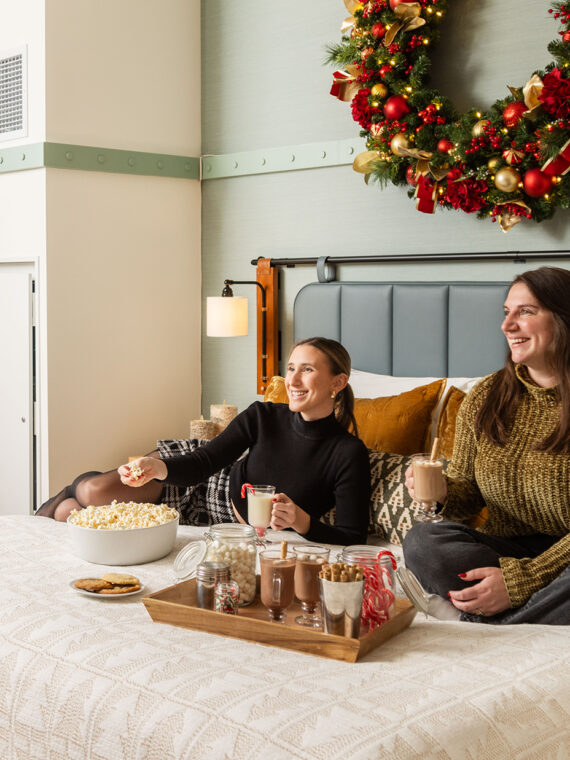Two people sit on a hotel bed while holding cups of hot chocolate. The hotel room is decorated with a large Christmas wreath and a Christmas tree.