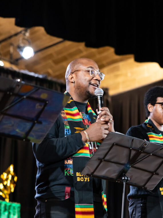 Four performers wearing colorful kente stoles sing into microphones on stage during a Kwanzaa celebration at the Penn Museum.