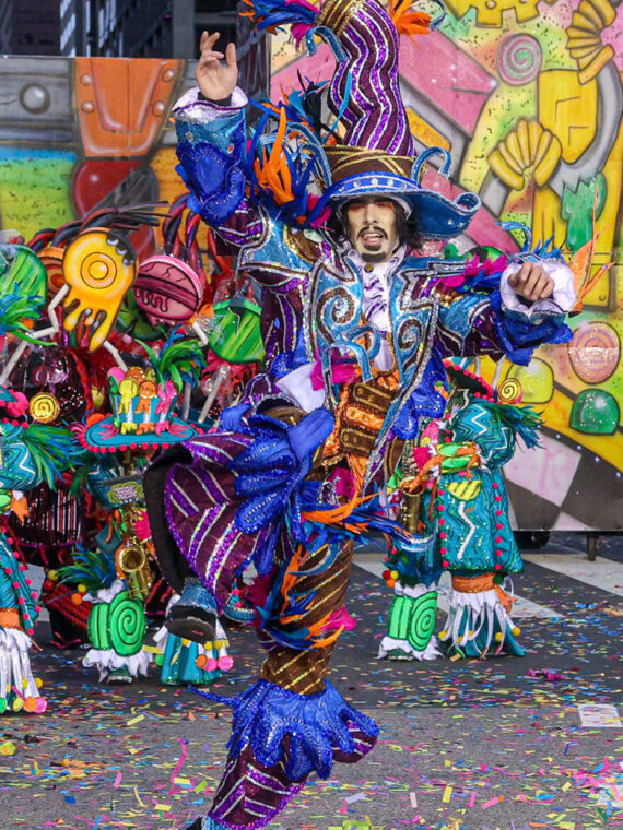 A guy dressed in very colorful attire leaping into the air while confetti falls and a band plays