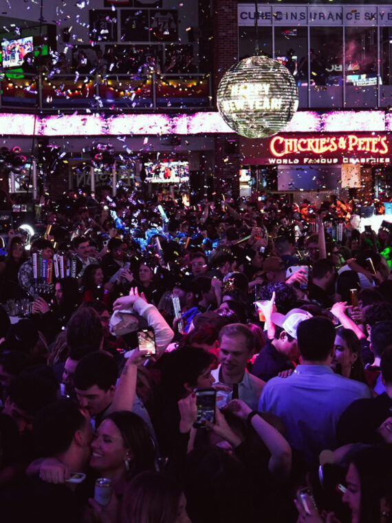 A packed New Year's Eve party with confetti falling, and glowing disco ball overhead and partygoers celebrating under glowing lights.